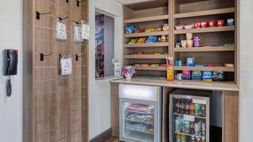Um pequeno quiosque de lanches e bebidas bem organizado, com várias prateleiras repletas de produtos diversos, uma pequena geladeira e um telefone na parede.