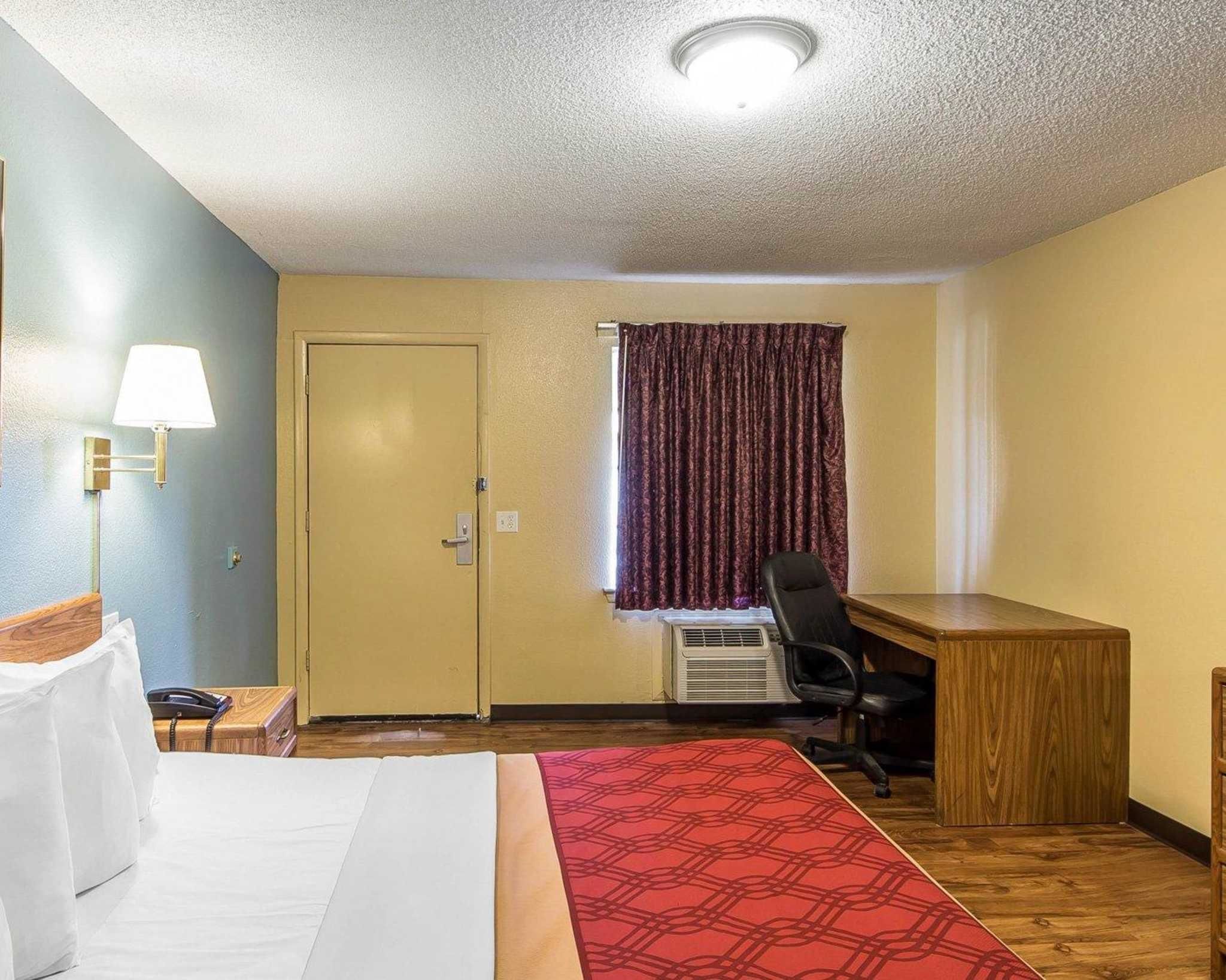 Quarto de hotel com cama, escrivaninha, cadeira, luminária e janela com cortinas.