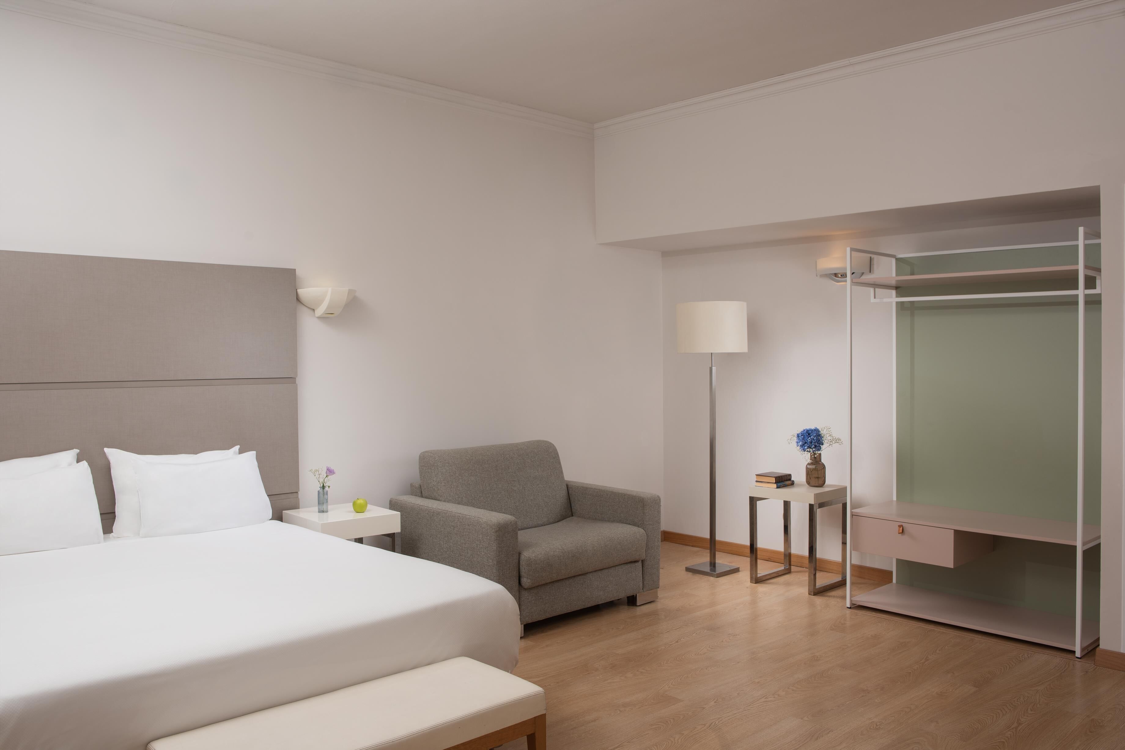 Quarto de hotel com cama, poltrona cinza, escrivaninha com abajur e guarda-roupa.