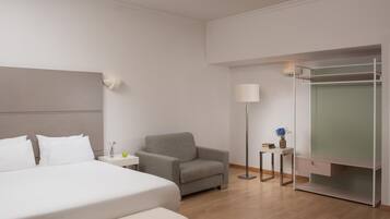 Quarto de hotel com cama, poltrona cinza, escrivaninha com abajur e guarda-roupa.