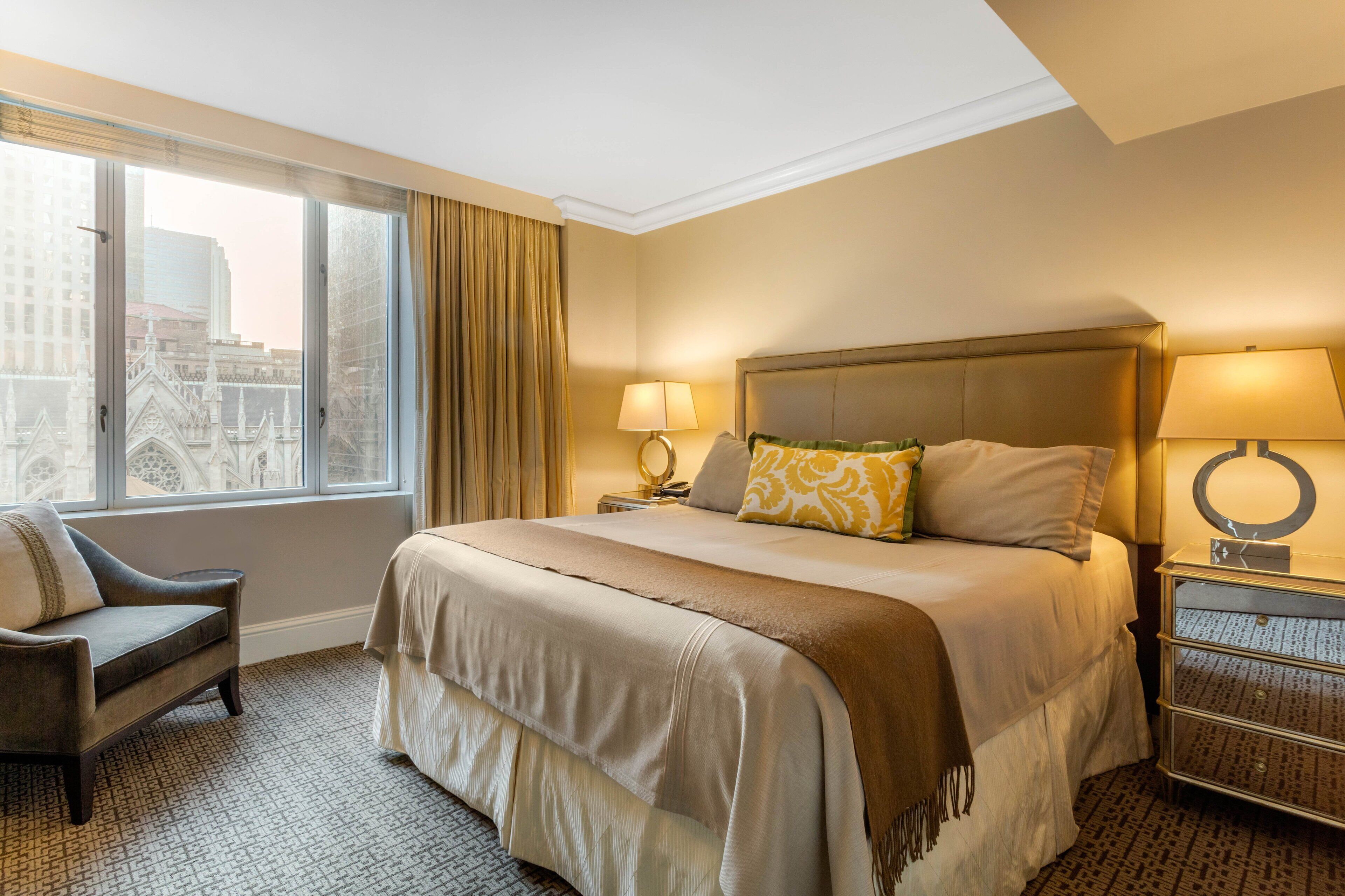Quarto de hotel com uma cama grande, uma cadeira, uma mesinha de cabeceira e vista para a cidade pela janela.