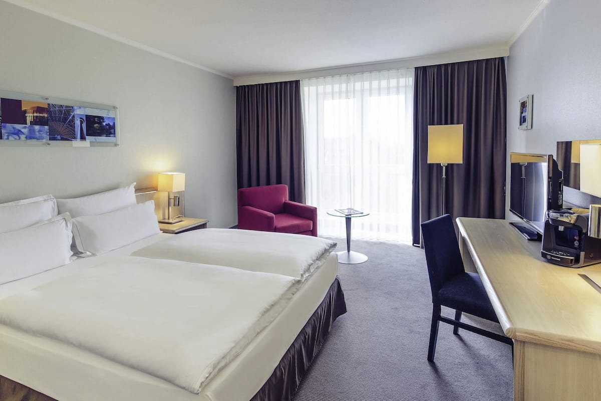 Quarto de hotel com uma cama grande, uma escrivaninha com computador, uma cadeira, uma mesinha e uma televisão.