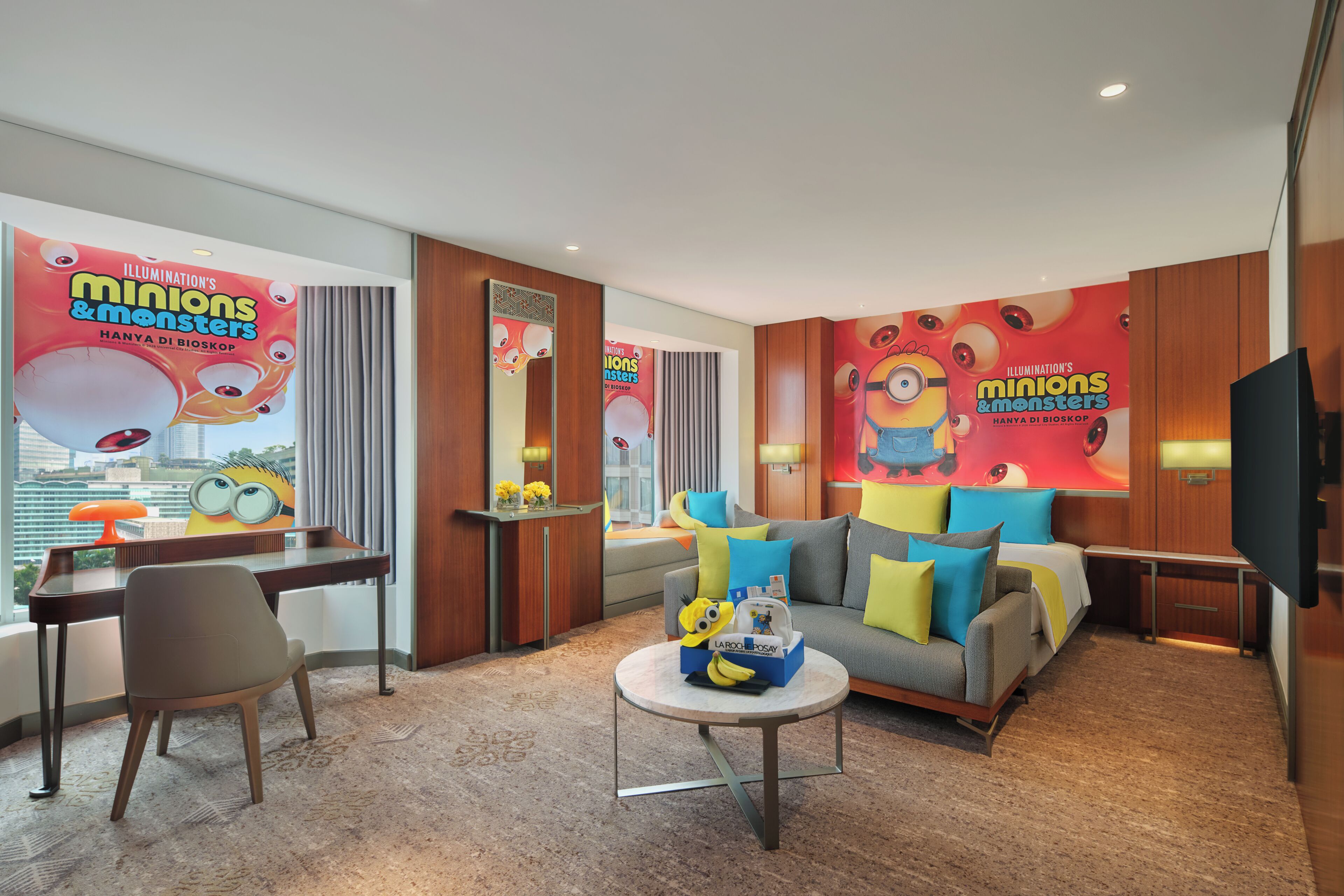 Quarto de hotel com decoração inspirada nos Minions, um sofá, uma mesa de trabalho e uma televisão.
