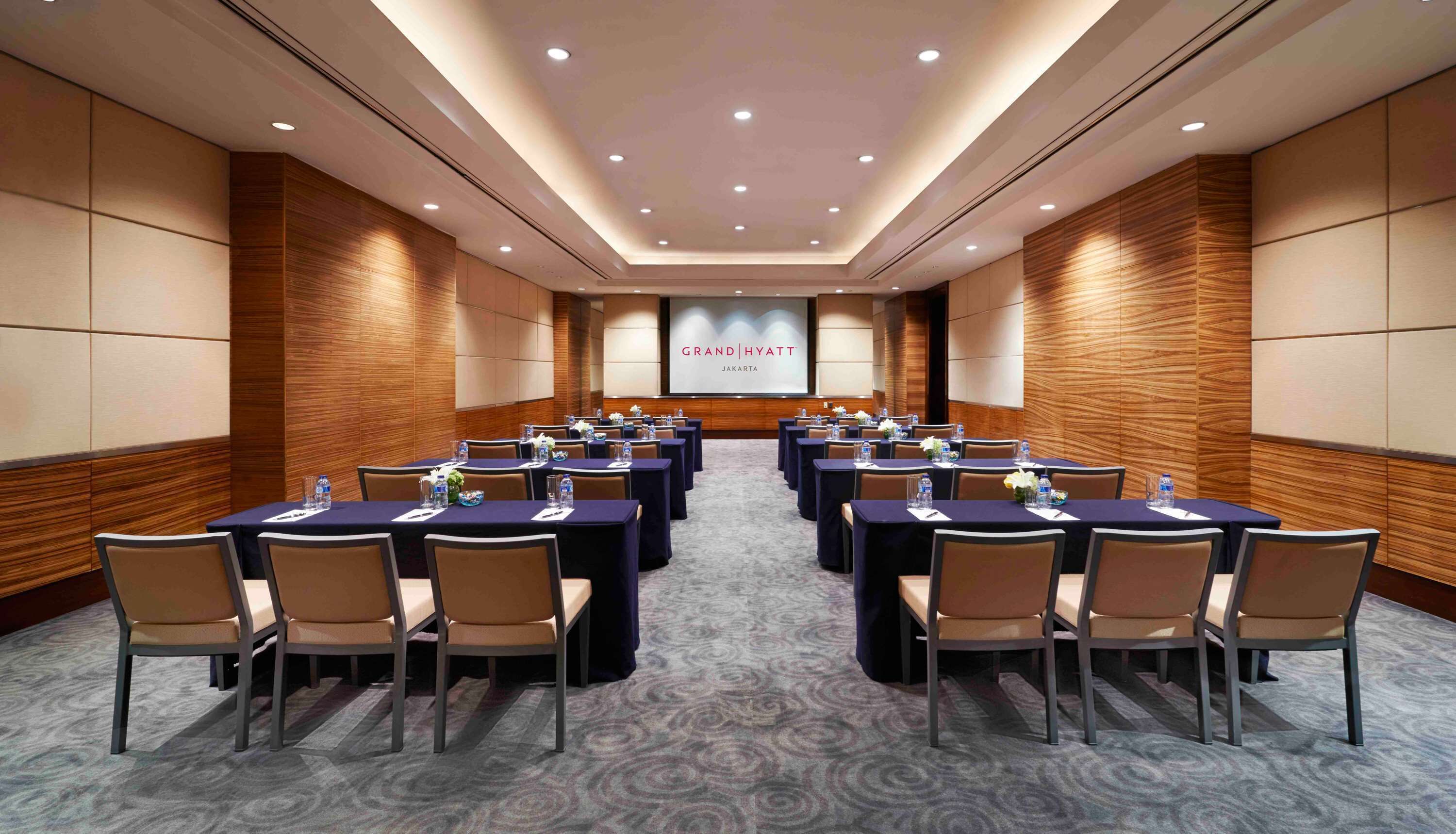 Uma sala de conferências com várias mesas e cadeiras, uma grande tela exibindo 'Grand Hyatt Jakarta' e piso coberto por carpete.