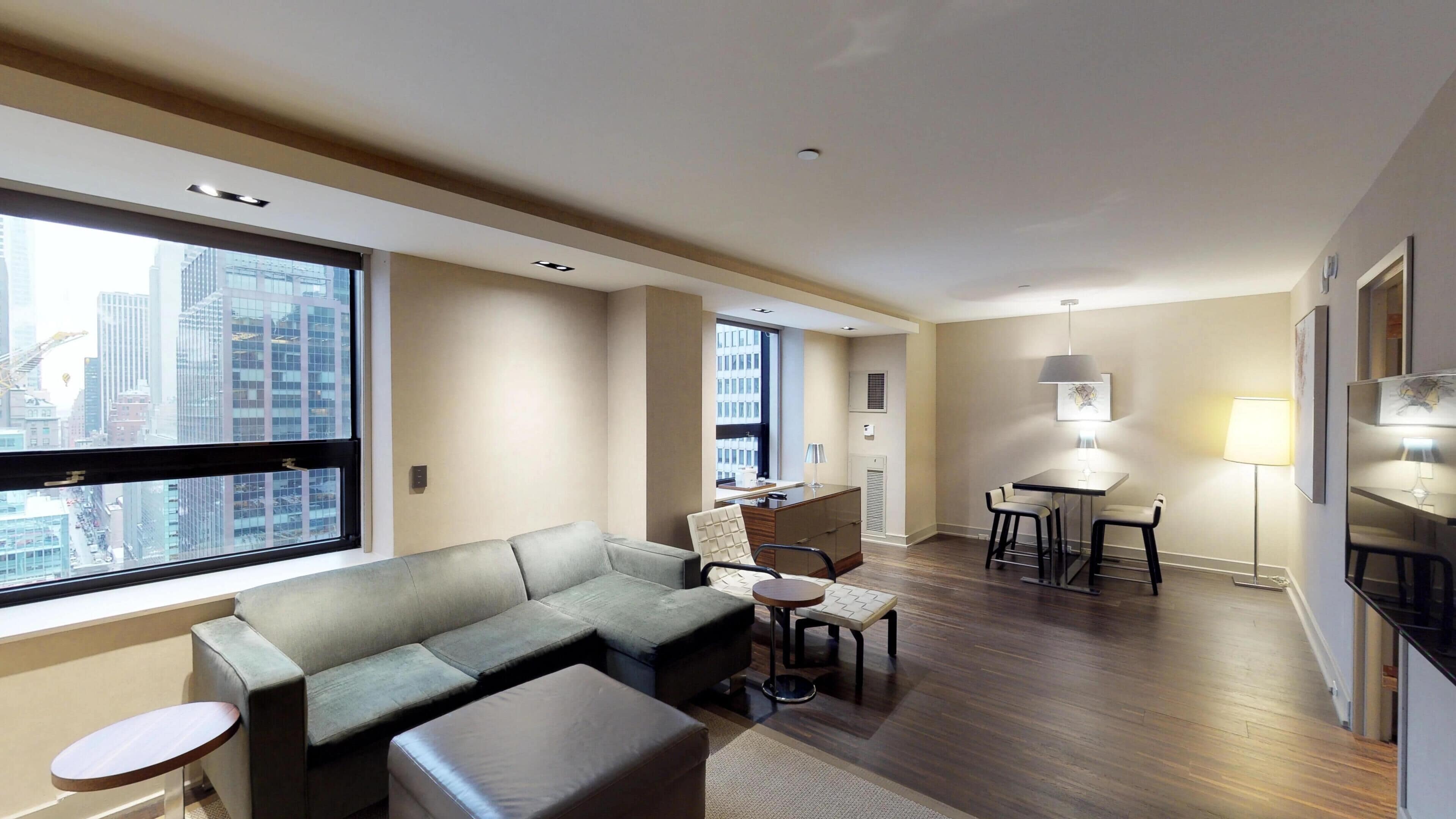 Uma sala de estar moderna com um sofá seccional, uma área de jantar com mesa e cadeiras, e janelas amplas com vista para a cidade.
