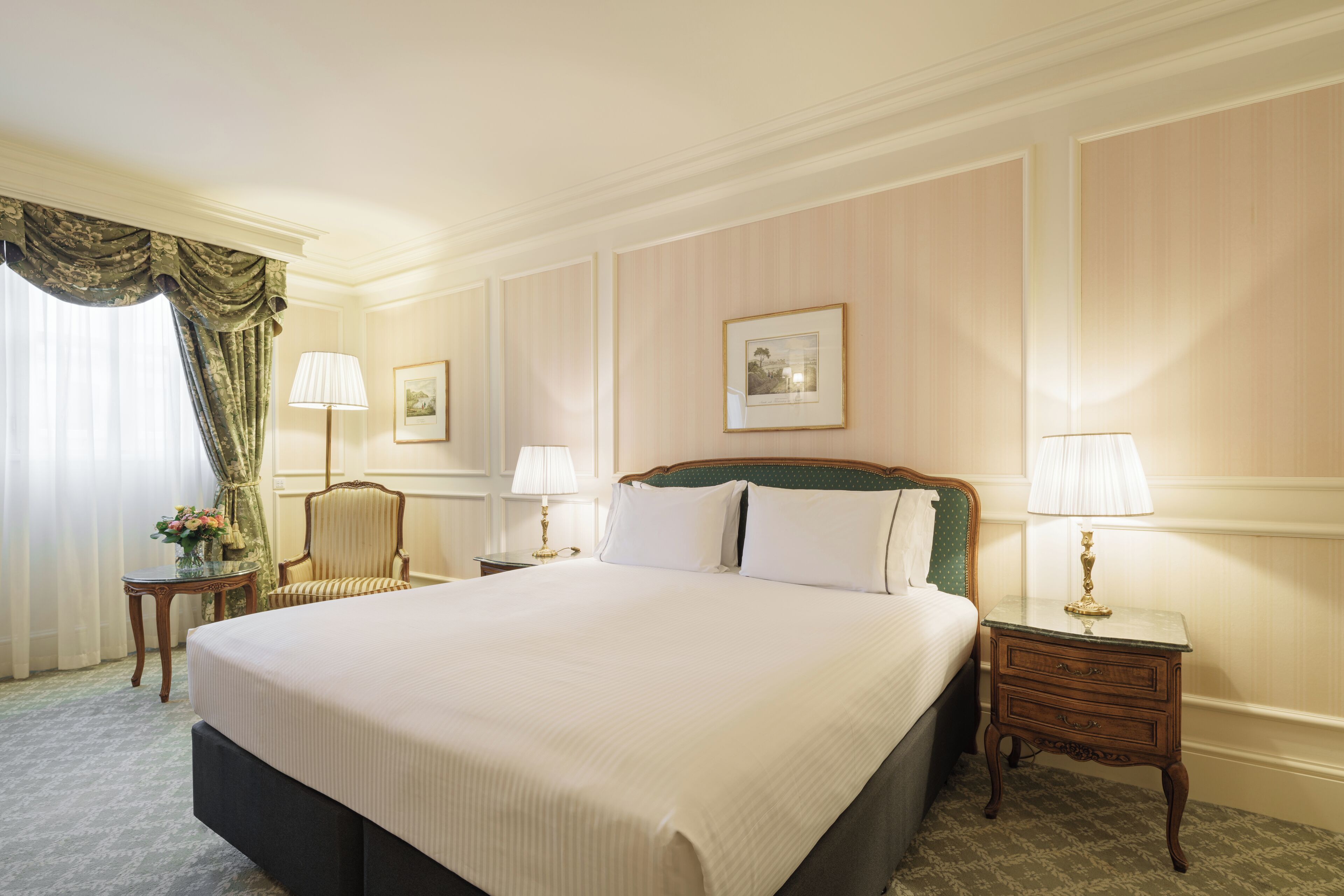 Quarto de hotel com uma cama grande, duas mesas de cabeceira com abajures, uma cadeira e um quadro na parede.