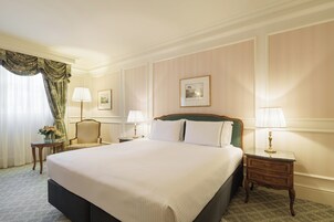 Quarto de hotel com uma cama grande, duas mesas de cabeceira com abajures, uma cadeira e um quadro na parede.