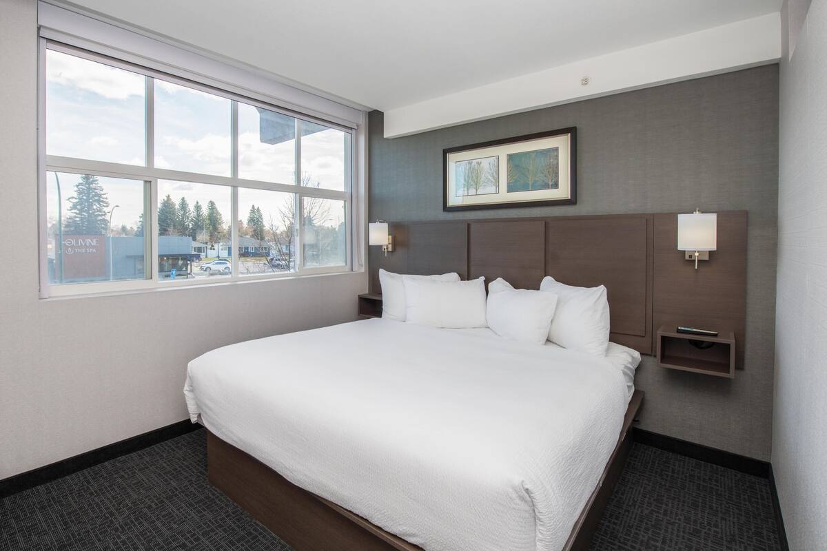 Quarto de hotel com uma cama grande, janela com vista para o exterior, quadro na parede e mesa de cabeceira com abajur.