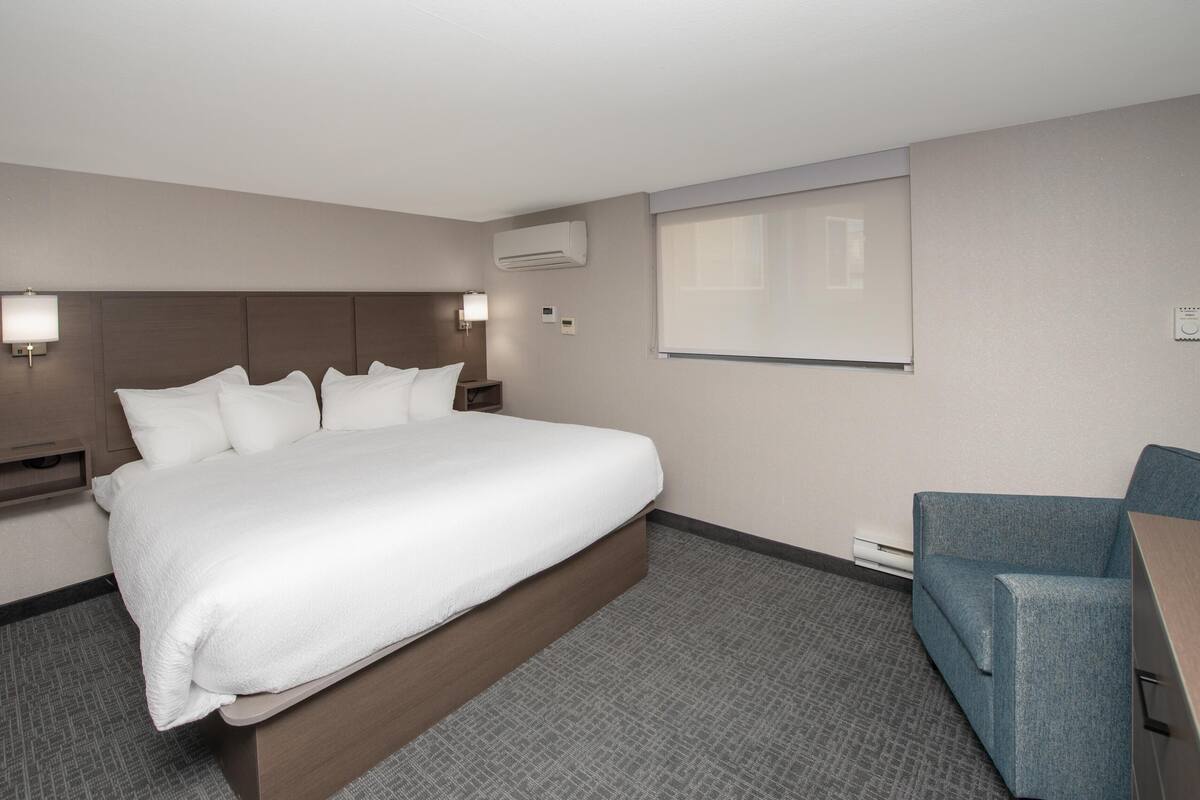 Quarto de hotel com uma cama grande, uma poltrona azul, ar condicionado fixado na parede e uma TV de tela plana.