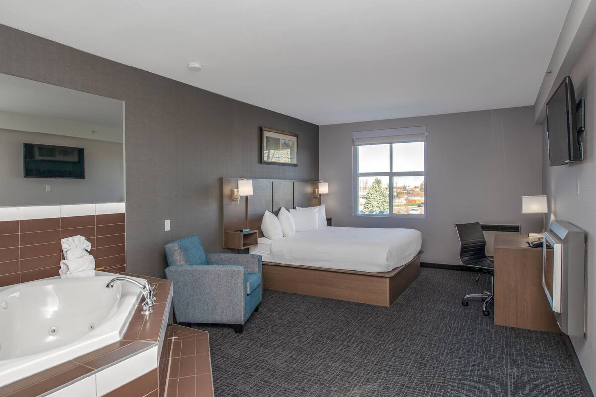Quarto de hotel com uma cama grande, área de estar com uma poltrona, escrivaninha, televisão e banheira.