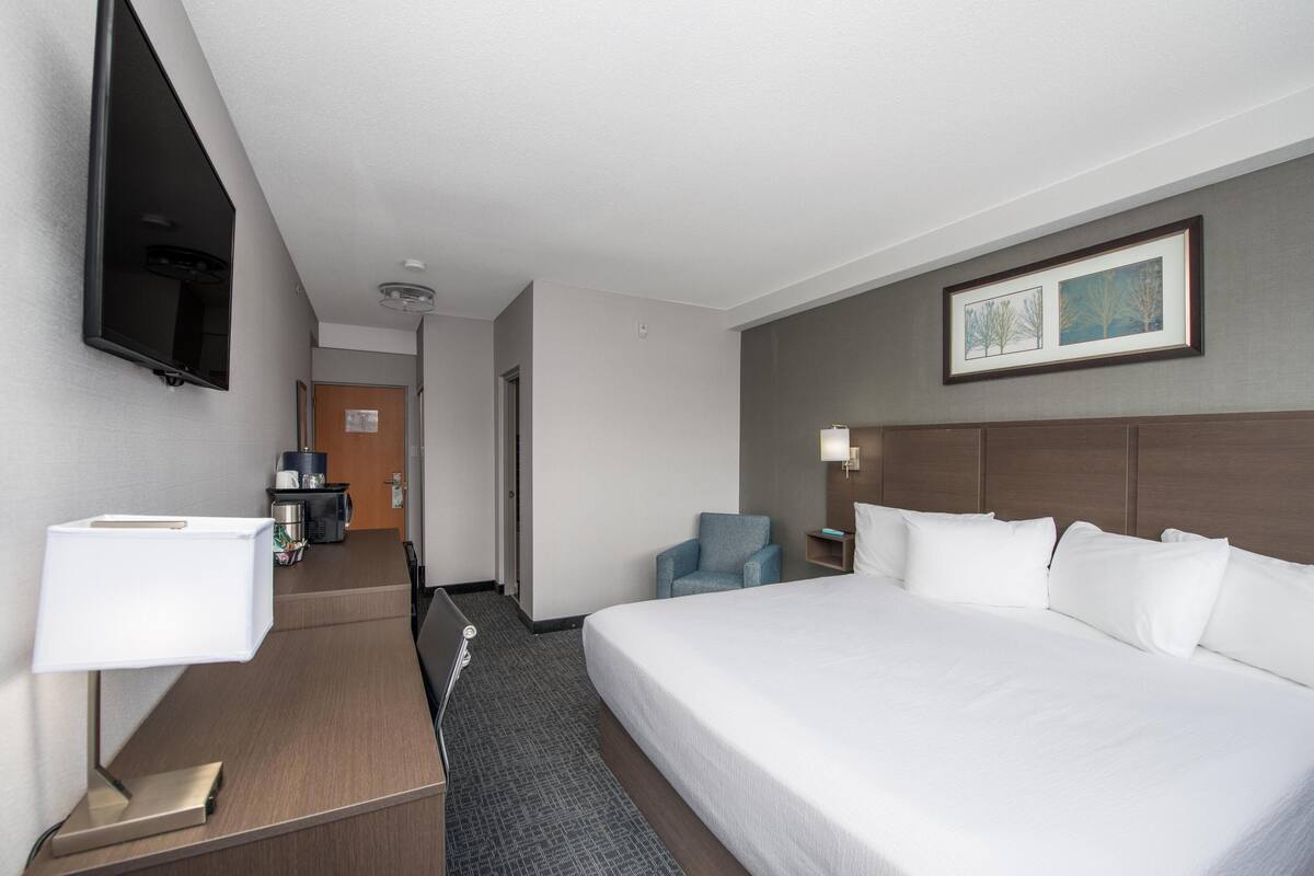 Quarto de hotel com uma cama, uma mesa de trabalho com luminária, uma cadeira, uma TV fixada na parede e um quadro emoldurado acima da cama.