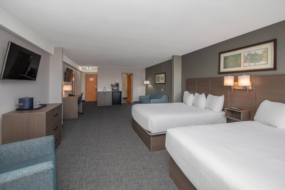 Quarto de hotel com duas camas, uma escrivaninha, uma cadeira, televisão, cozinha compacta e banheiro.