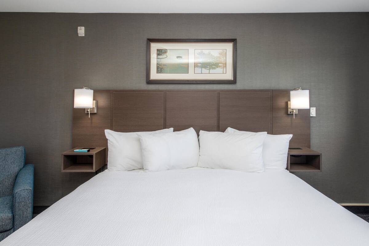Quarto de hotel com uma cama grande, duas mesas de cabeceira com abajures, um quadro na parede e uma cadeira azul.