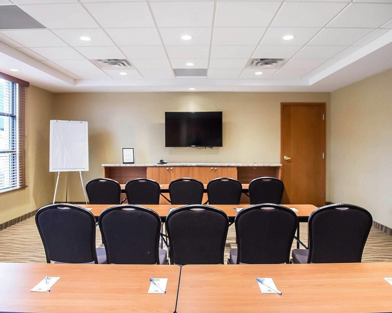 Uma sala de conferências com uma mesa comprida, cadeiras pretas, um quadro branco, uma televisão de tela plana e uma porta.