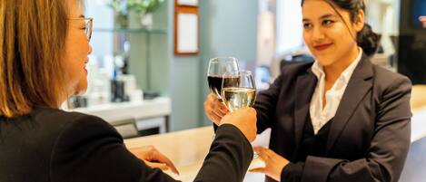 Duas pessoas sentadas à mesa, cada uma segurando um copo de vinho. O ambiente parece ser um espaço interno com design moderno, com divisória de vidro e paleta de cores neutras.