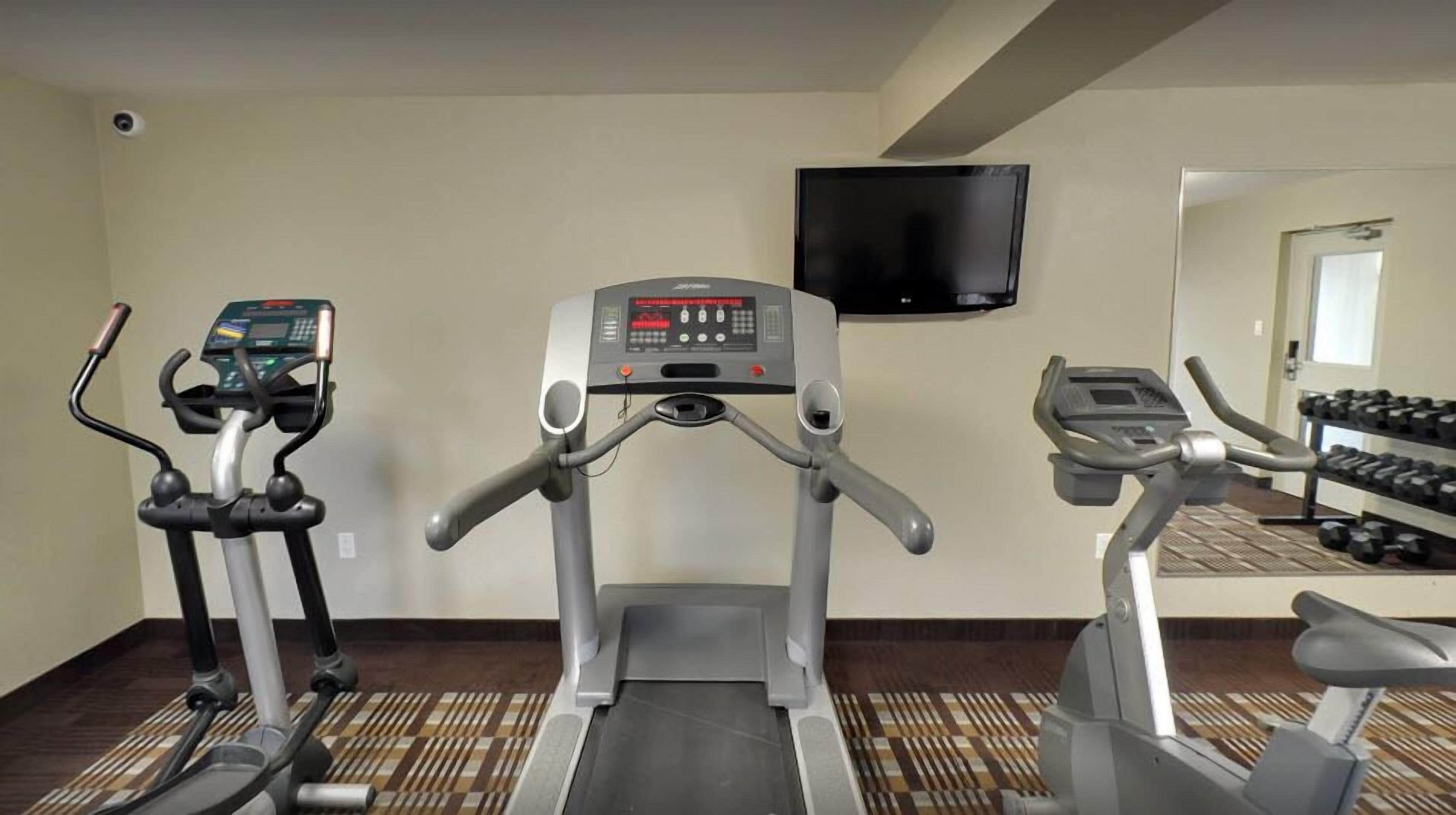Três bicicletas ergométricas numa sala com televisão de tela plana na parede e um suporte com halteres.