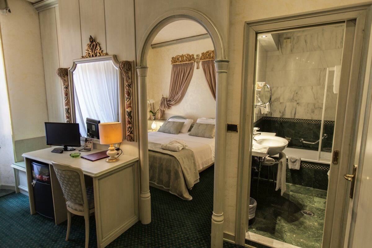 Quarto de hotel com escrivaninha, computador, luminária, cama e banheiro com pia e espelho.