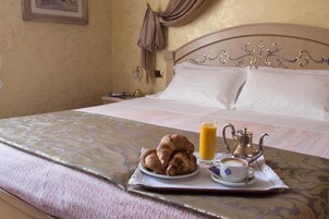 Cama bem arrumada com cabeceira decorativa, bandeja com croissants, copo de suco de laranja e bule de chá com xícara de café sobre toalha de mesa estampada.