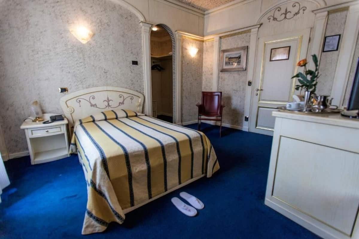 Quarto de hotel com cama, mesa de cabeceira, cadeira, escrivaninha com espelho e porta.