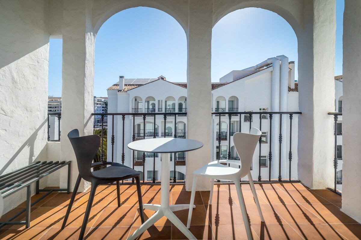 Varanda com duas cadeiras e uma mesa pequena, com vista para um edifício com janelas em arco.