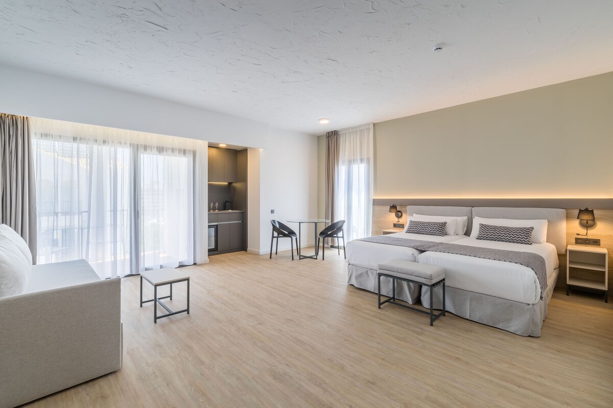 Quarto de hotel moderno com uma cama grande, um sofá, uma mesa pequena e uma escrivaninha. Possui uma kitchenette no canto e vista para a cidade através de grandes janelas.