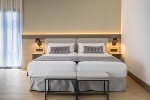 Uma cama de casal com um banco cinzento, duas mesas de cabeceira com luminárias e uma cabeceira em tom claro.