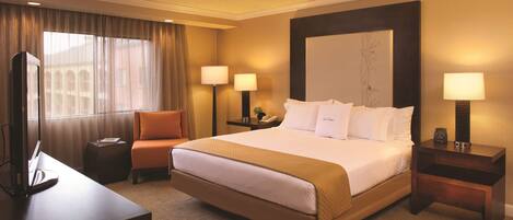 Quarto de hotel com uma cama grande, duas mesas de cabeceira, uma escrivaninha, uma cadeira e uma televisão.
