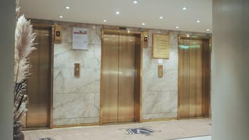 Quatro elevadores com paredes de mármore e detalhes dourados, chão coberto por carpete com um logotipo e uma placa com instruções de emergência.