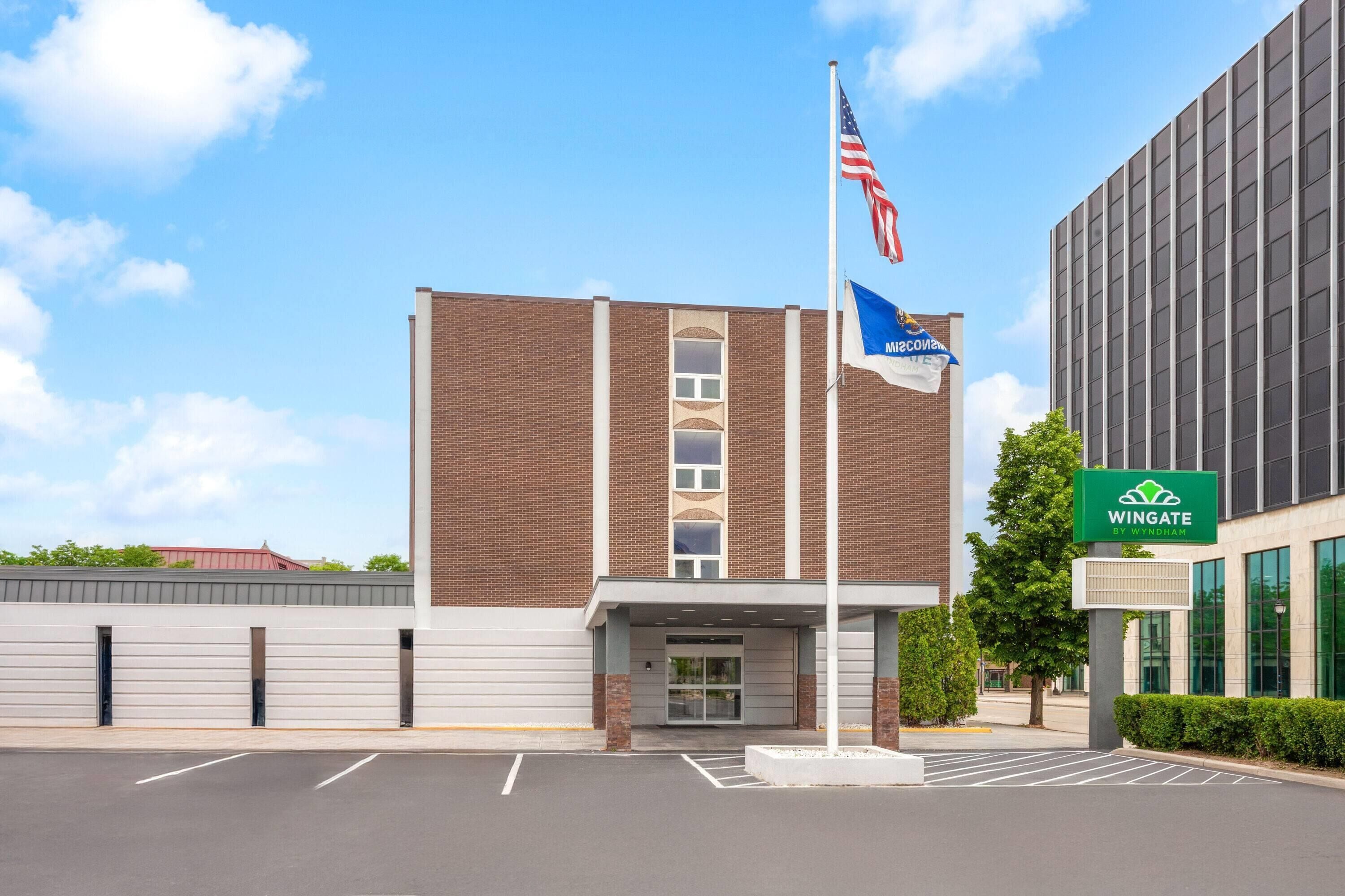 Um edifício com fachada de tijolos, um mastro com a bandeira americana e uma placa indicando 'WINGATE BY WYNDHAM'.