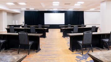 Uma sala de conferências com uma tela grande, várias mesas e cadeiras dispostas para reuniões.