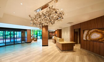 Um saguão de hotel moderno com um grande lustre, balcão de recepção em madeira e vista para o exterior.