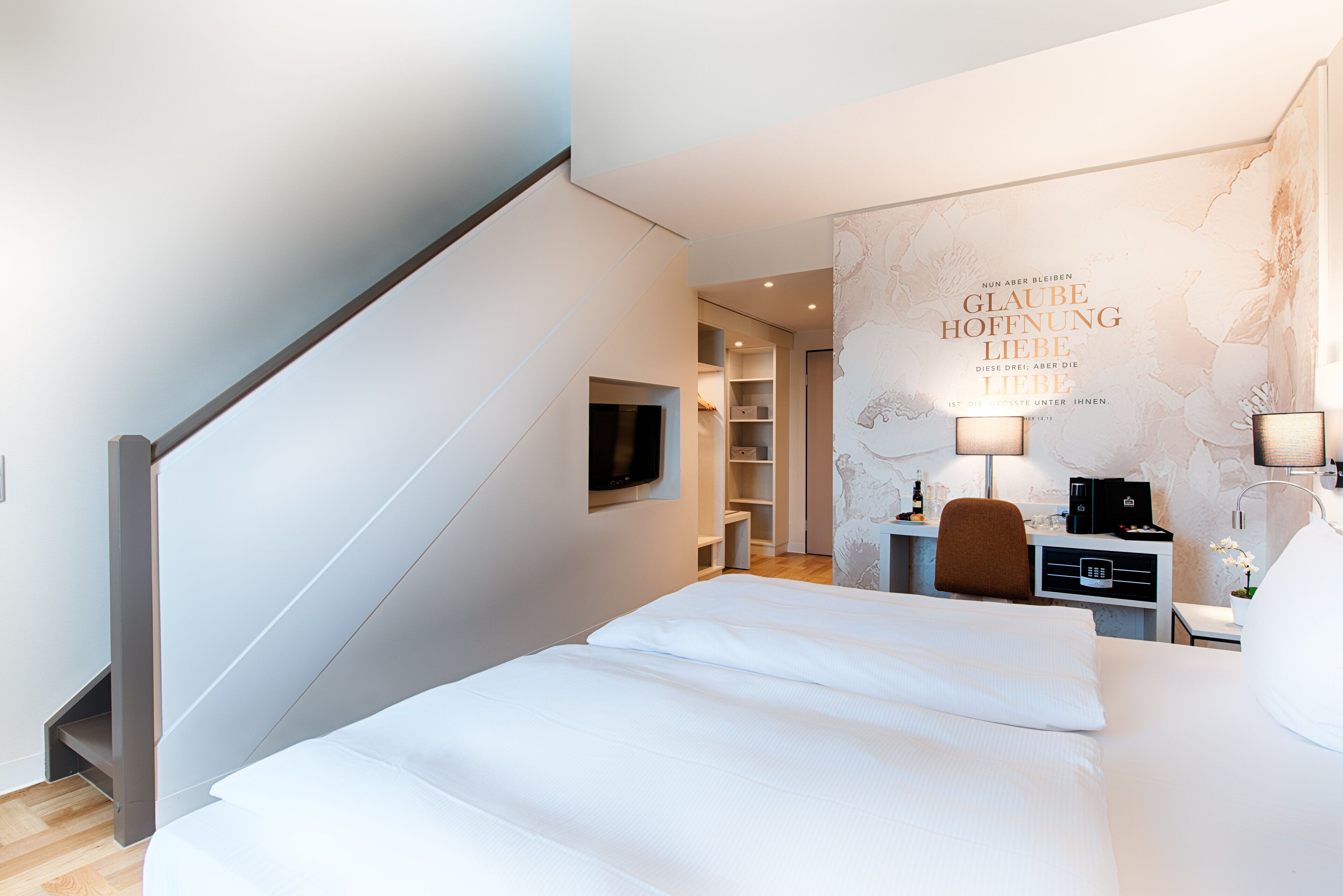 Quarto de hotel moderno com uma cama grande, uma escrivaninha com computador e uma TV fixada na parede.