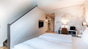 Quarto de hotel moderno com uma cama grande, uma escrivaninha com computador e uma TV fixada na parede.
