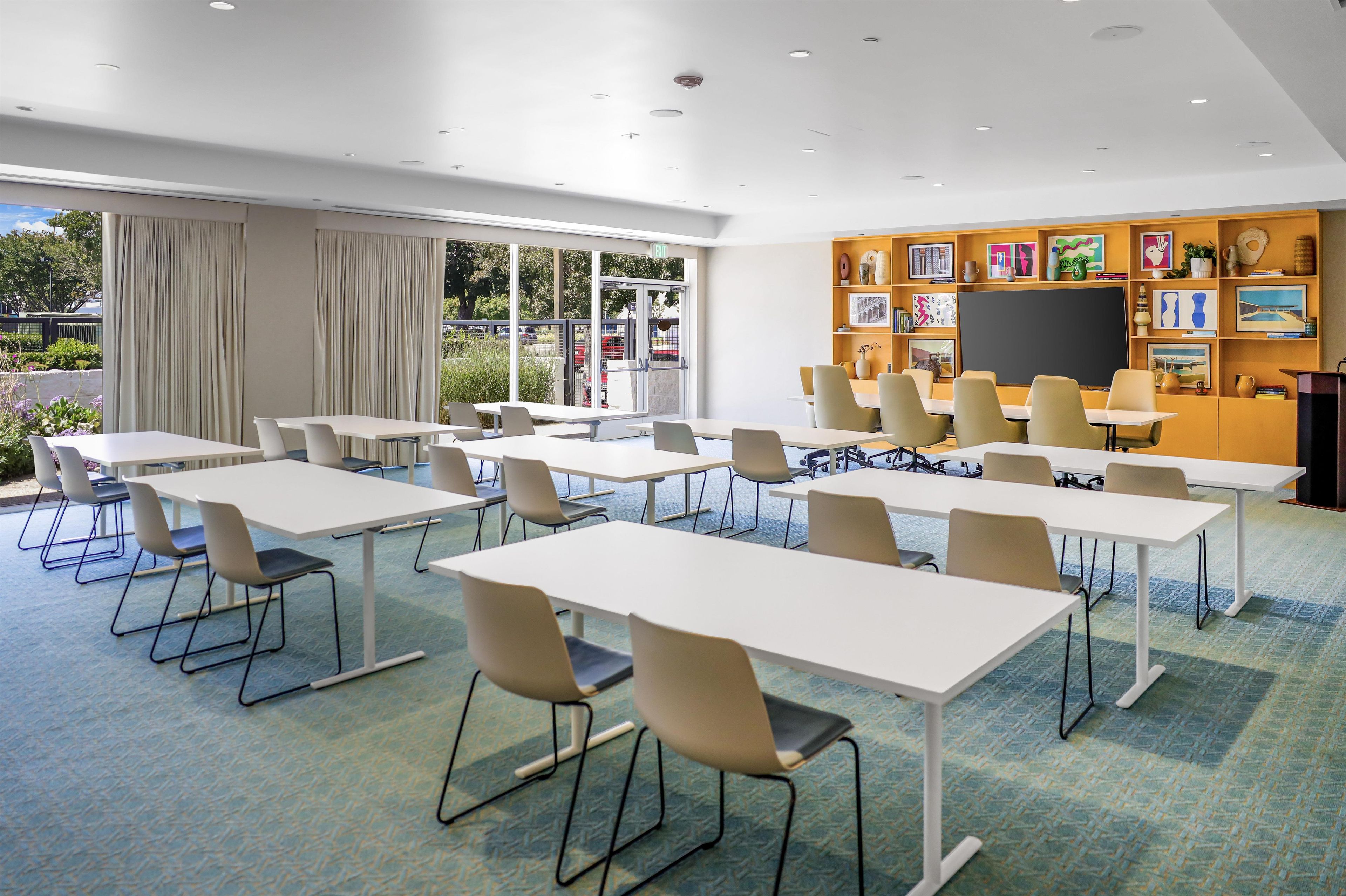 Uma sala de aula com mesas, cadeiras, um quadro negro e uma estante com diversos objetos.