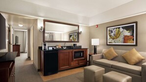 Quarto de hotel com cozinha compacta, sofá, mesa de centro, televisão e uma pintura grande na parede.