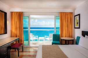 Quarto de hotel com cama, escrivaninha, cadeira, mesa pequena e vista para o oceano através de grandes portas de vidro.