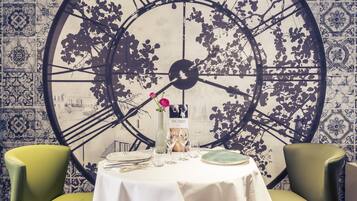 Uma mesa de jantar posta com pratos, copos e um vaso de flores, com um relógio grande e azulejos estampados na parede.