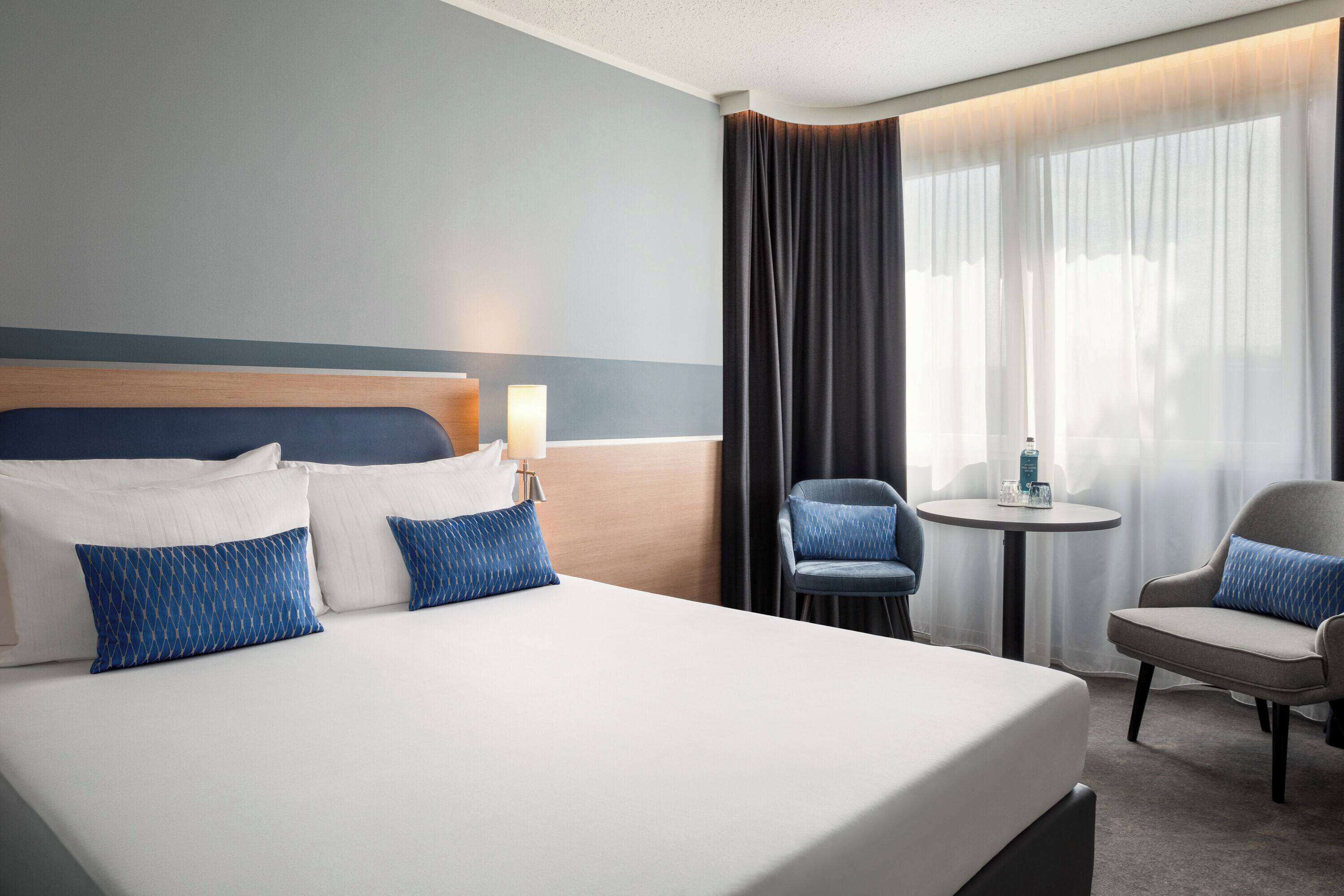 Quarto de hotel com uma cama grande, duas cadeiras, uma mesa pequena e uma janela com cortinas.