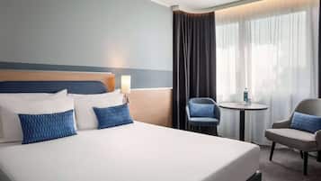 Quarto de hotel com uma cama grande, duas cadeiras, uma mesa pequena e uma janela com cortinas.