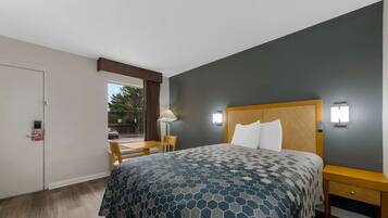 Quarto de hotel com uma cama grande, uma mesinha de cabeceira, uma escrivaninha e uma janela com cortinas.
