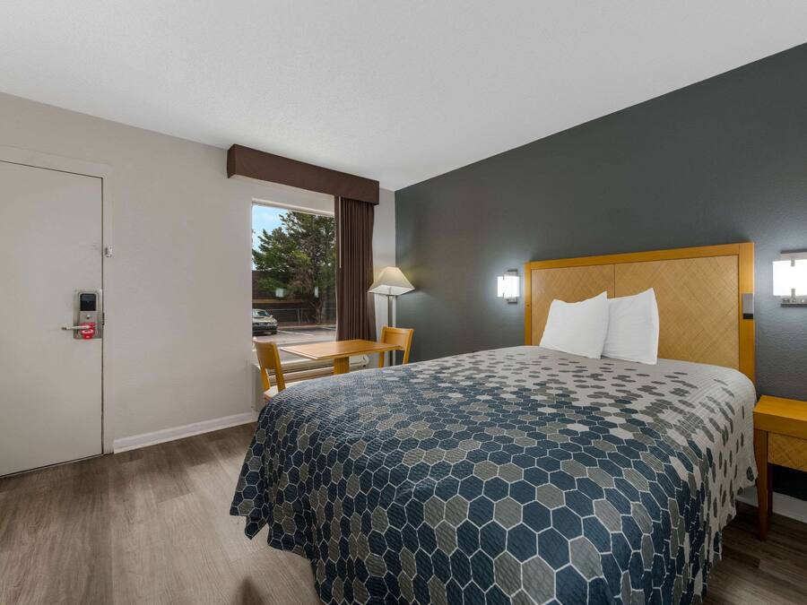 Quarto de hotel com uma cama grande, uma mesinha de cabeceira, uma escrivaninha e uma janela com cortinas.