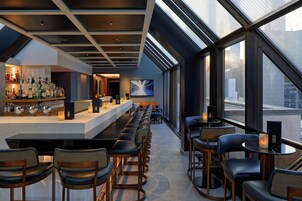 Uma área de bar moderna com um balcão comprido, banquetas e vista para a cidade através de grandes janelas.