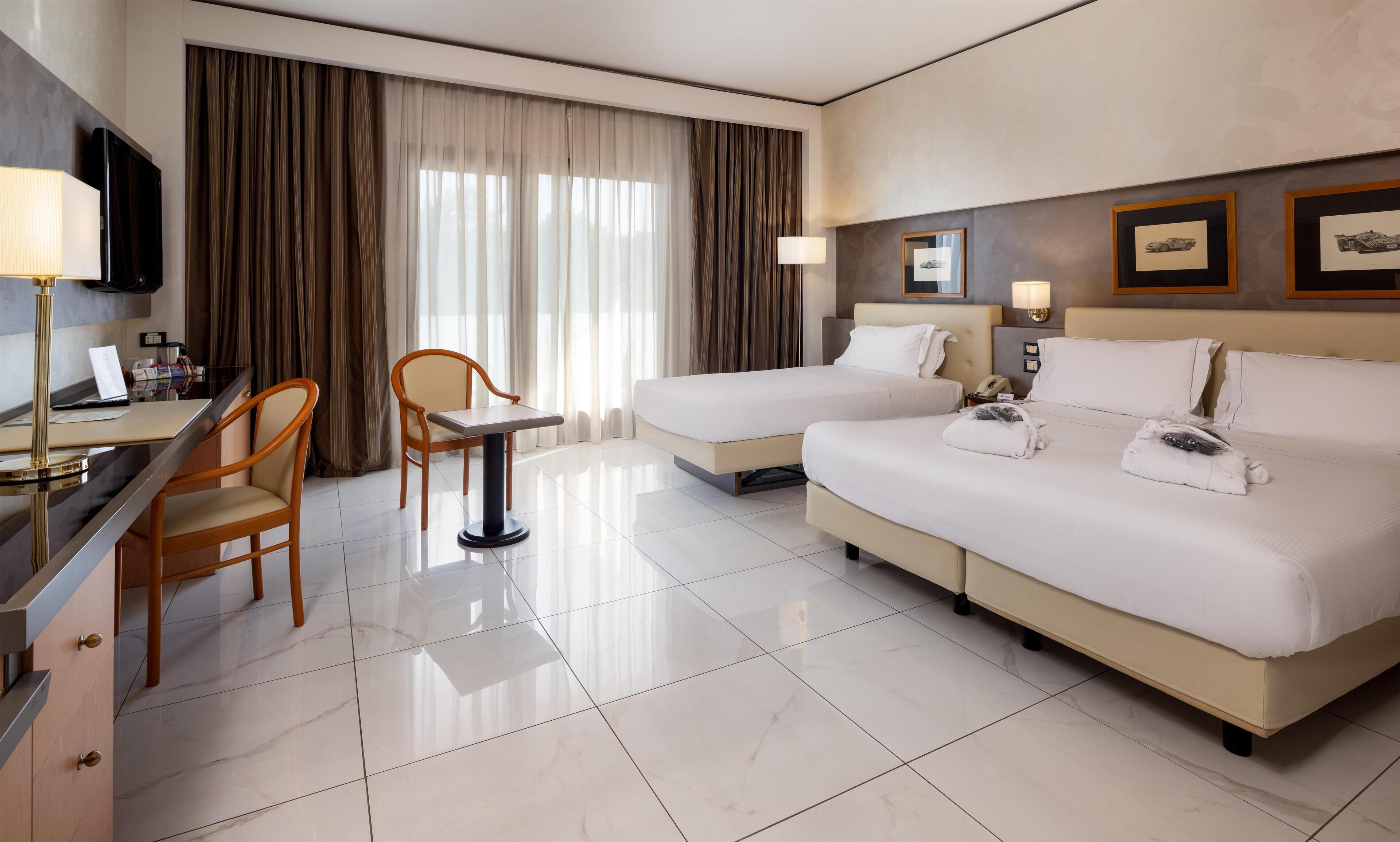 Quarto de hotel com uma cama, duas mesas de cabeceira, uma escrivaninha com cadeira, uma televisão e cortinas.