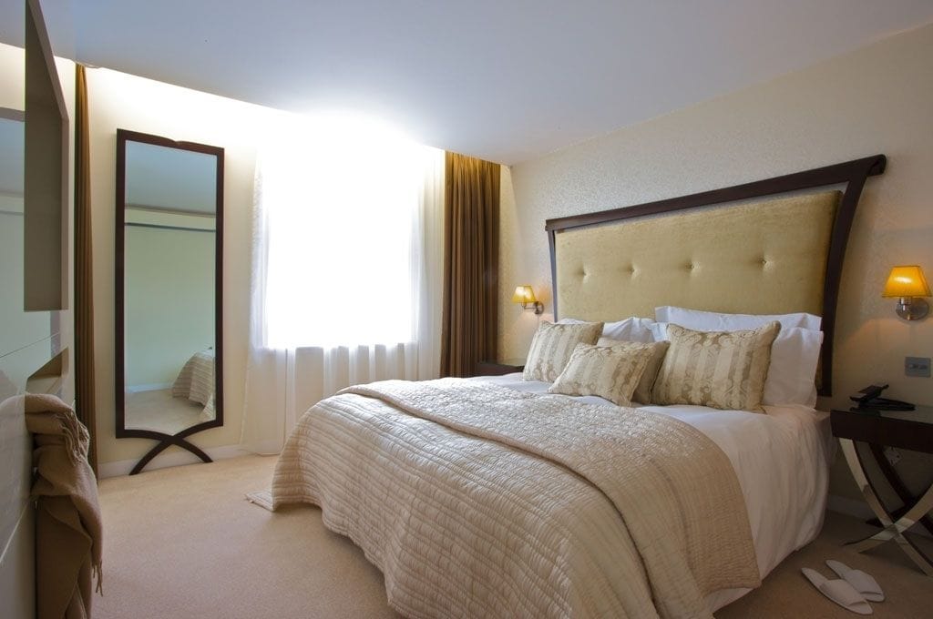 Quarto de hotel com uma cama grande, um espelho, uma mesa de cabeceira com abajur e vista para o exterior.