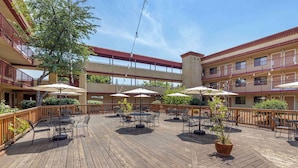 Um deck de madeira com assentos externos e guarda-sóis, cercado por um edifício de vários andares com sacadas.