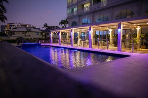 Uma área de piscina com um espaço de estar coberto e um edifício ao fundo.