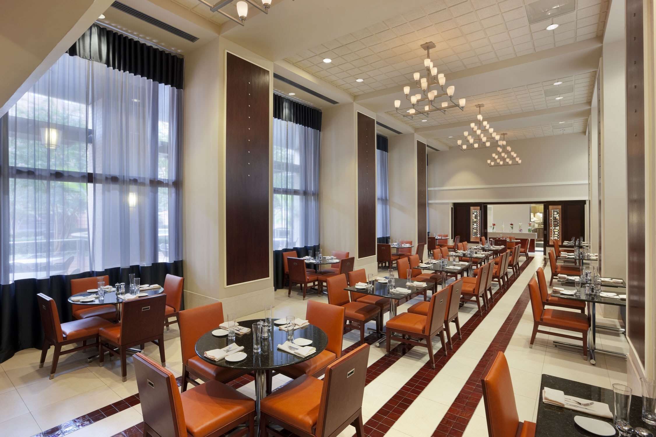 Uma ampla sala de jantar de hotel, com várias mesas e cadeiras, janelas grandes com cortinas leves e um lustre moderno.