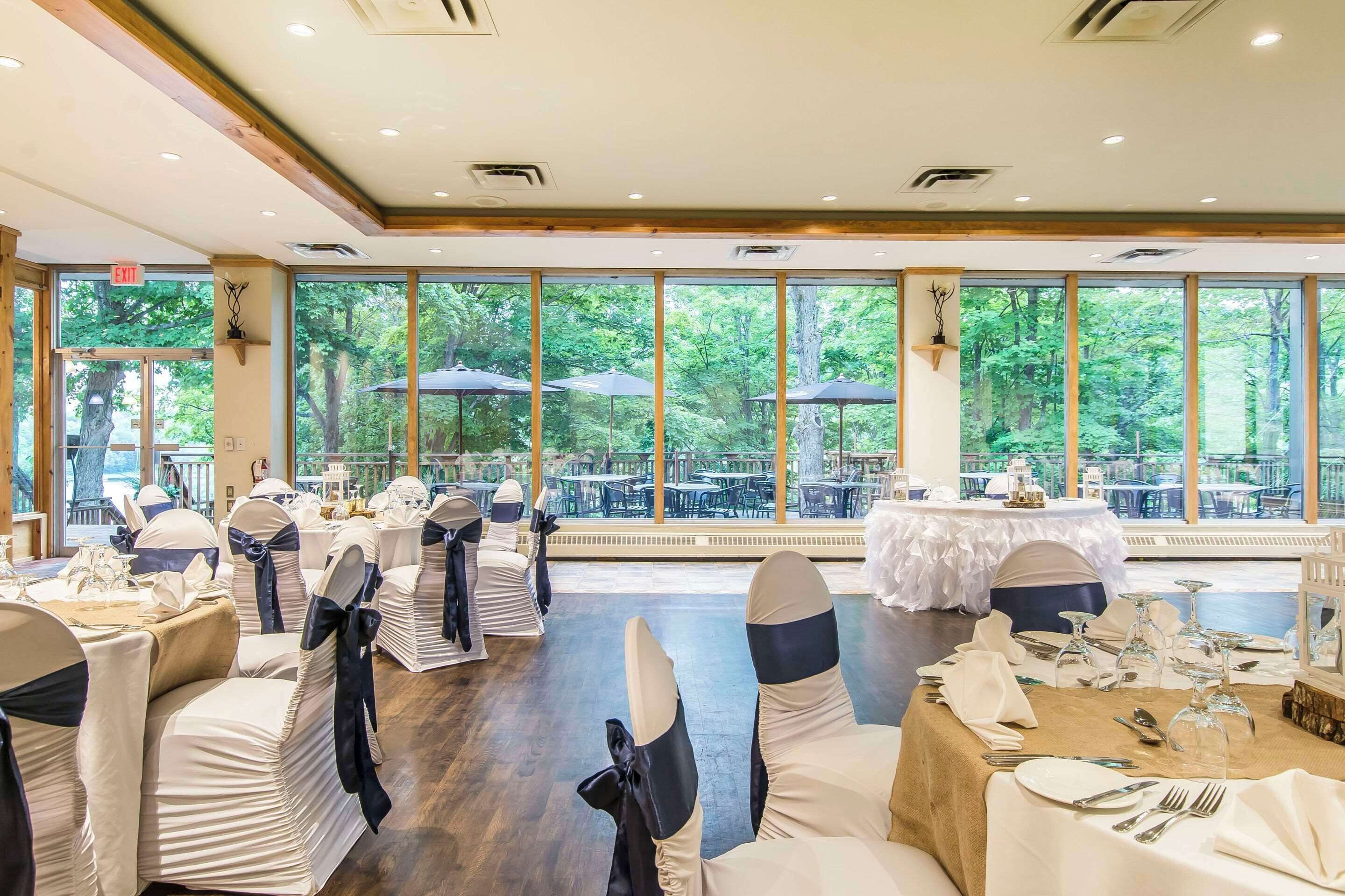 Uma sala de banquetes com mesas redondas preparadas para refeições, grandes janelas com vista para uma floresta e um sinal de saída.