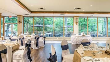 Uma sala de banquetes com mesas redondas preparadas para refeições, grandes janelas com vista para uma floresta e um sinal de saída.