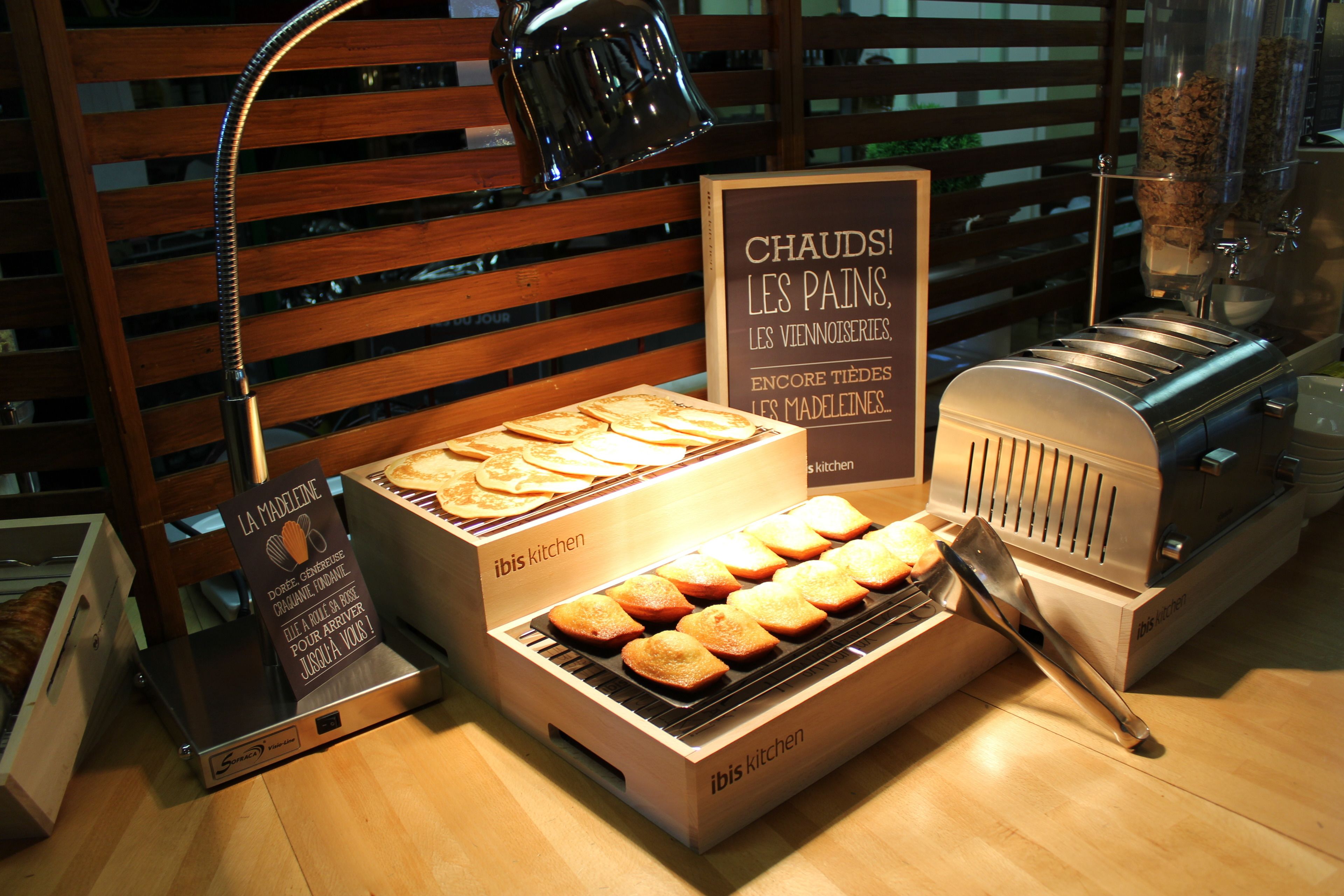 Vitrine de padaria com pão e doces recém-assados, um letreiro anunciando 'Chaud's Les Pains, Les Viennoiseries, Encore Tièdes Les Madeleines', e uma máquina de café.